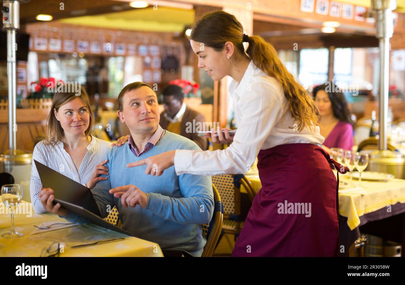 Girl waiter helps a couple choose dishes from the menu in restaurant ...