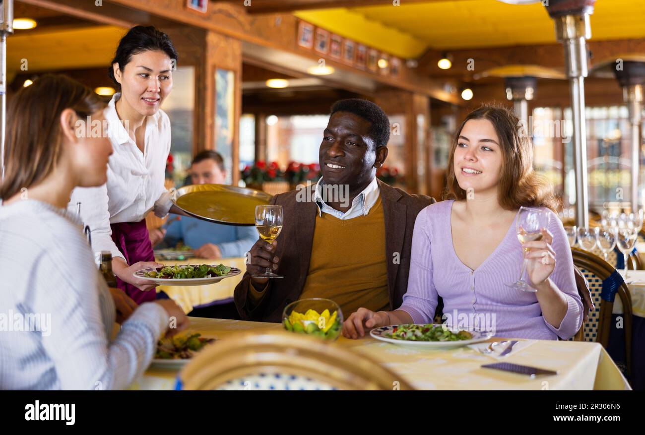 Asian waitress serving cheerful man and women over dinner in restaurant ...