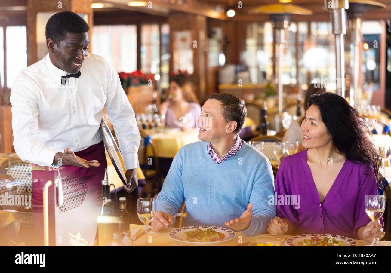Positive biracial couple talking to friendly African American waiter in ...