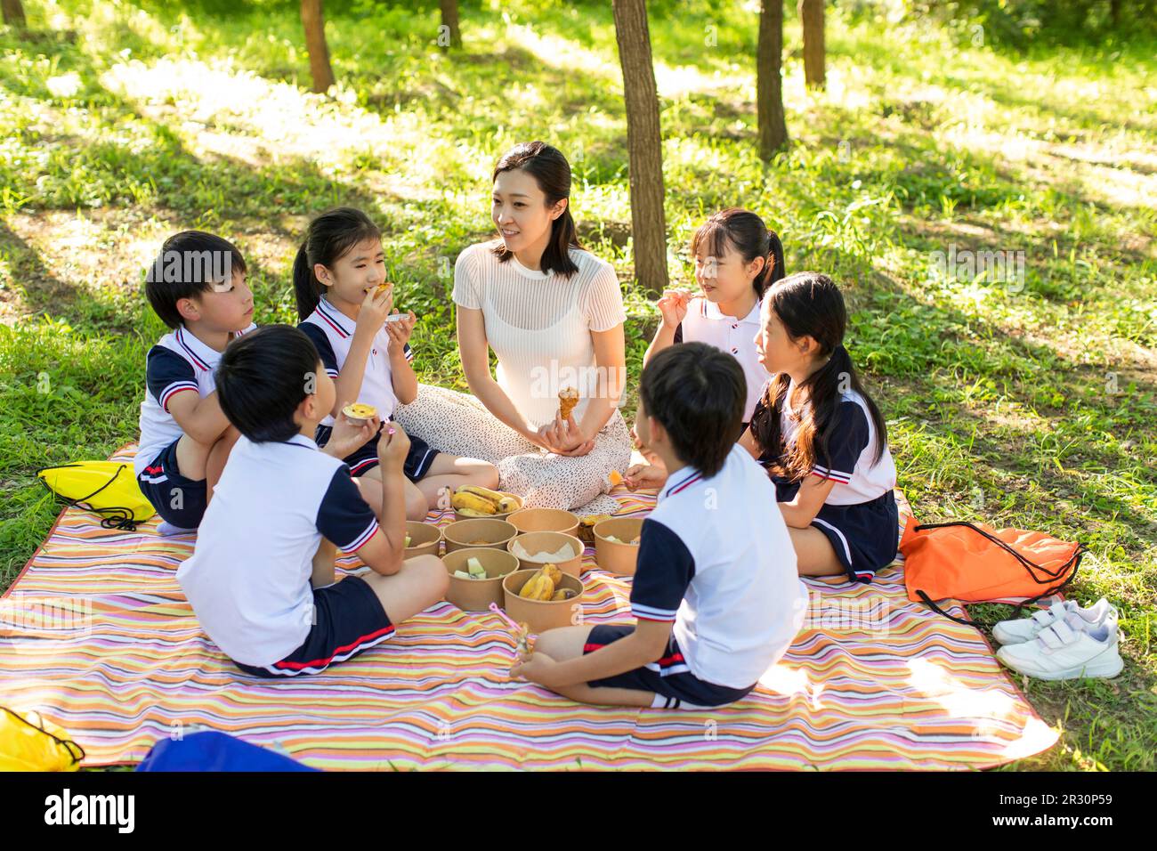 Cheerful Chinese school children picnicking outdoors with their teacher ...