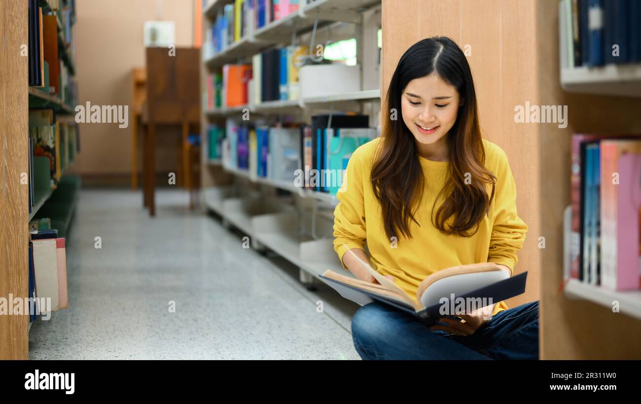 Concentrated young female sitting on floor near bookshelves, reading ...