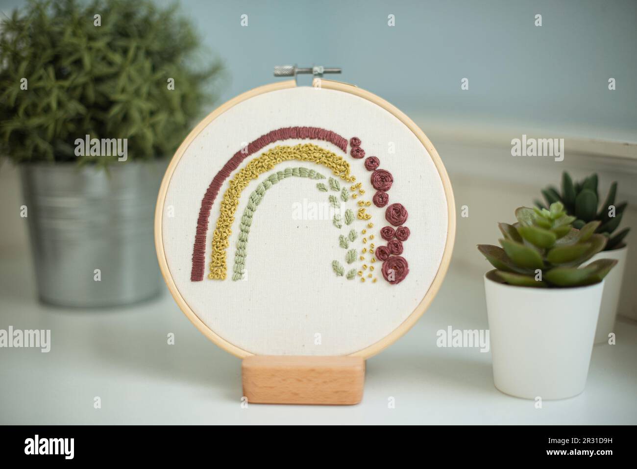 Homemade embroidery hoop on a sideboard next to assorted houseplants ...