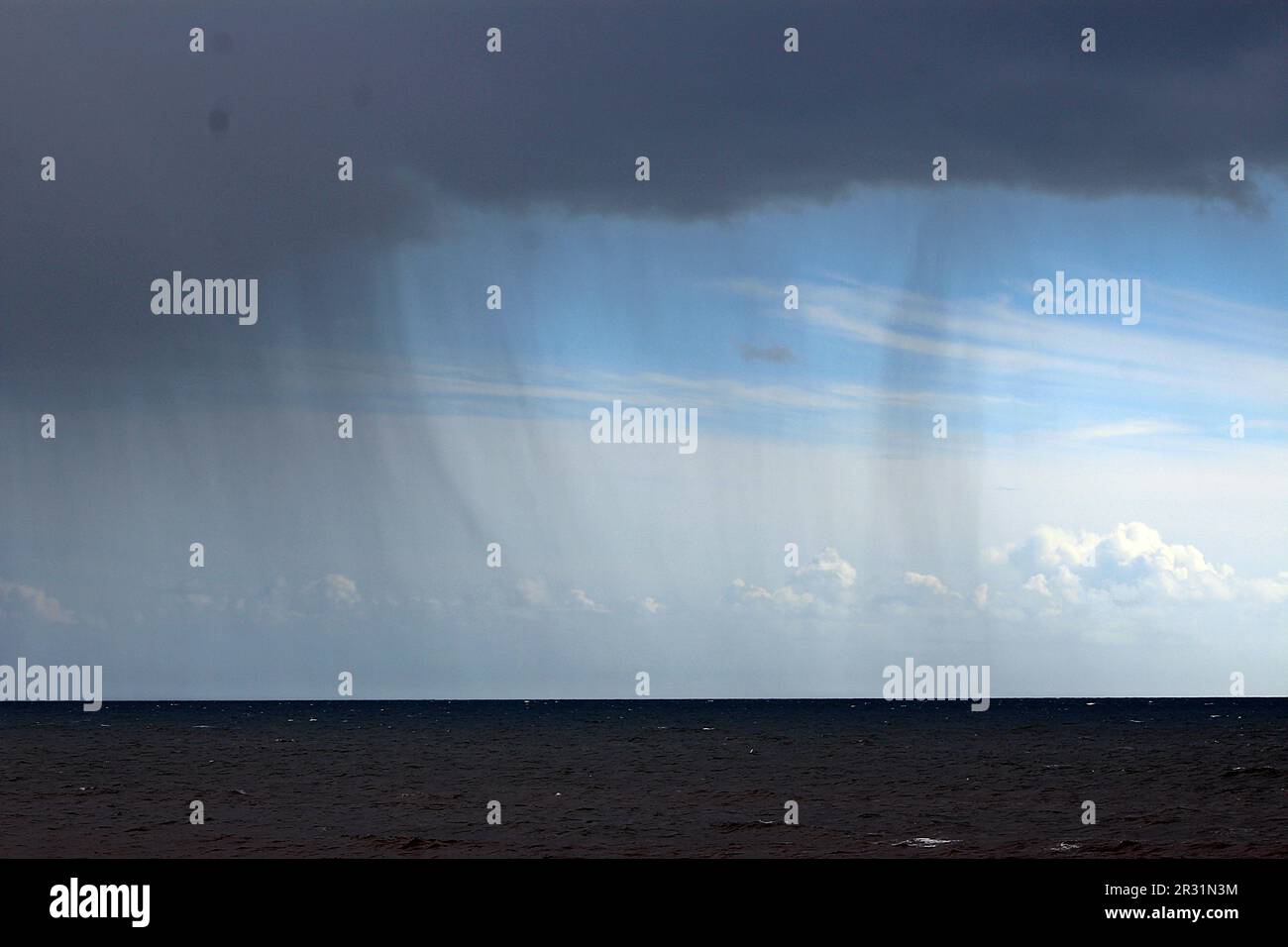 rain falling from storm clouds above a dark sea with blue sky in the ...
