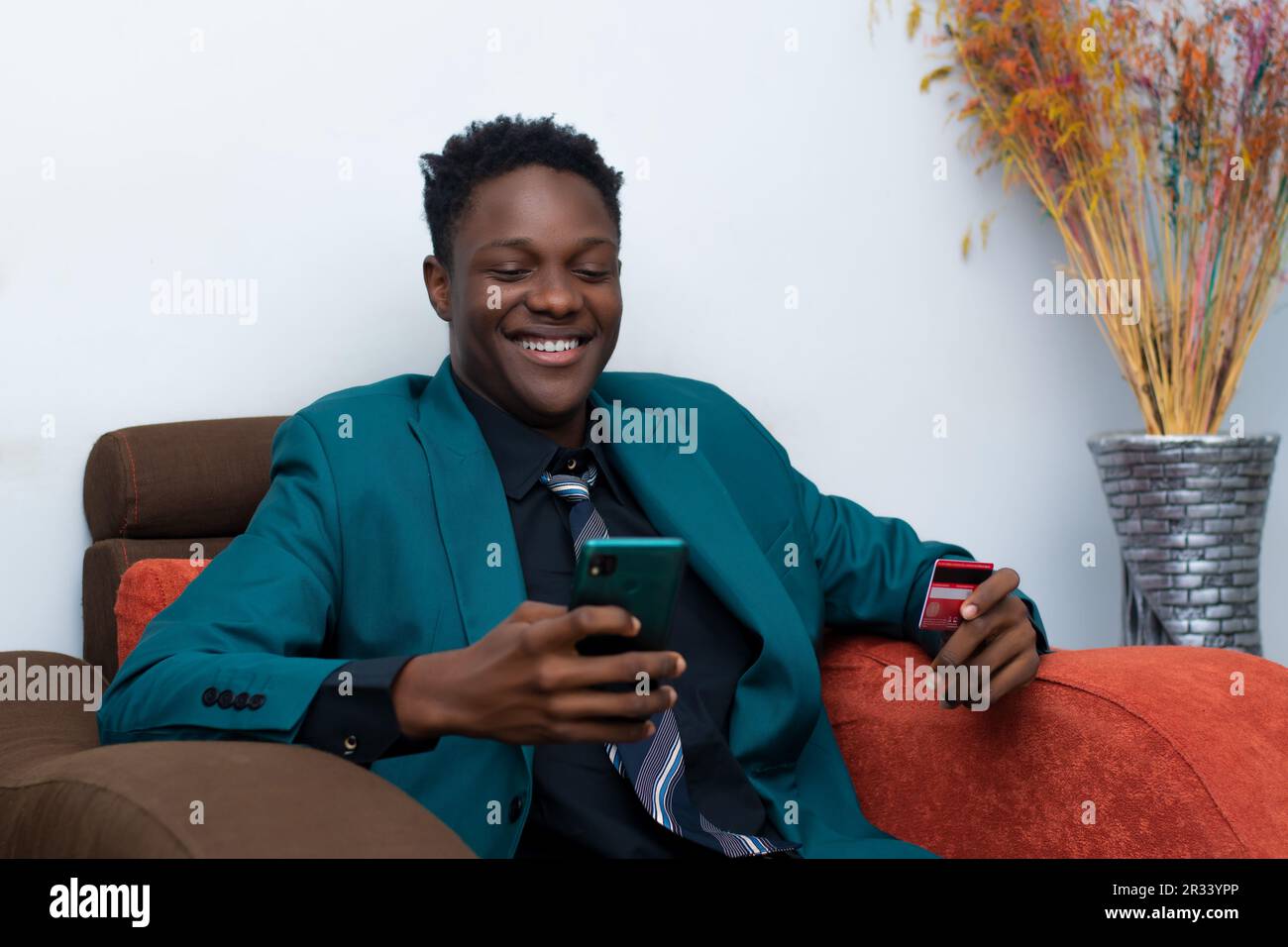 handsome young black man wearing a suit using his phone and credit card, sitting on a chair ...