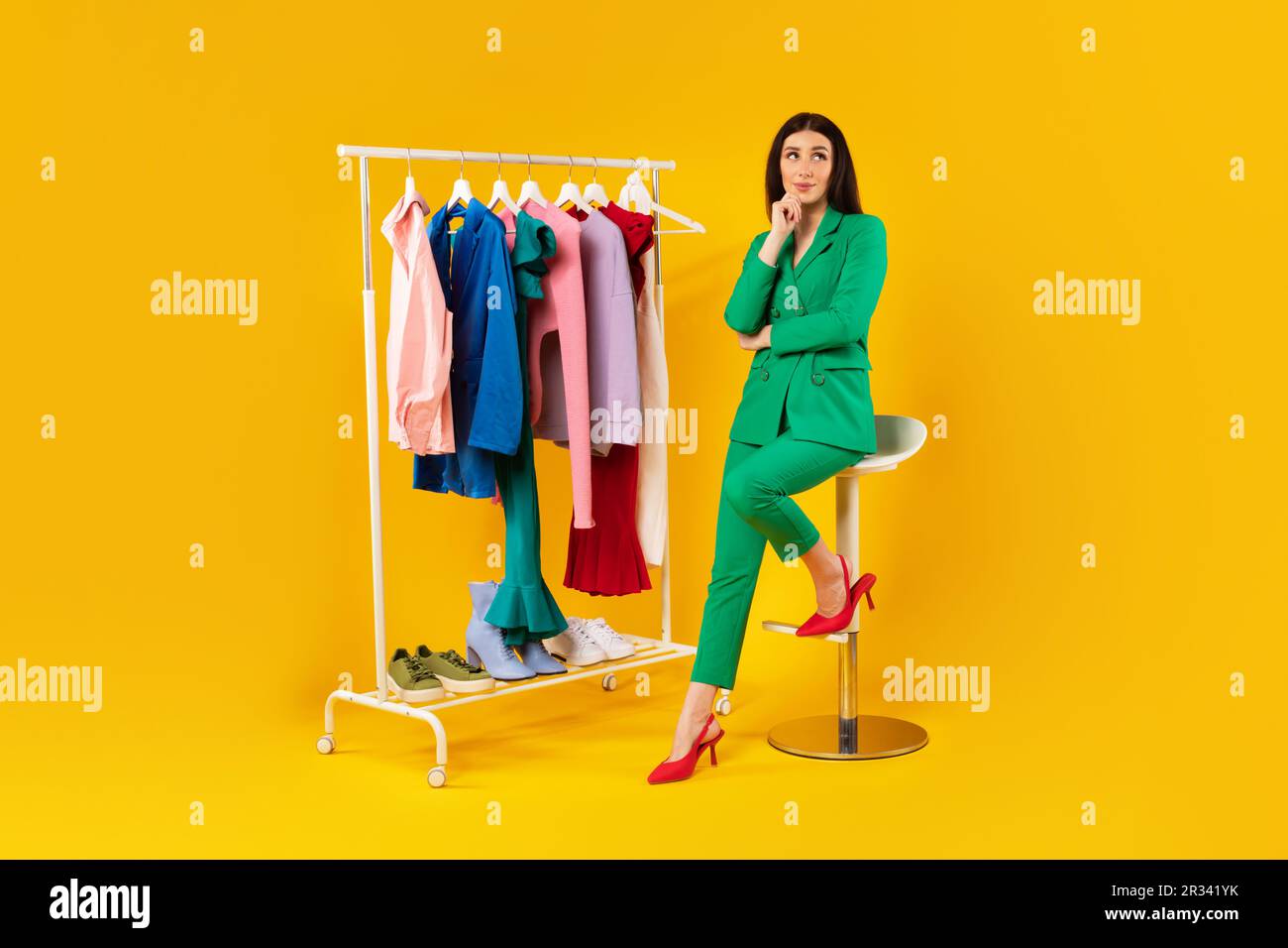 Pretty stylist lady sitting near garment rack and choosing new clothes ...