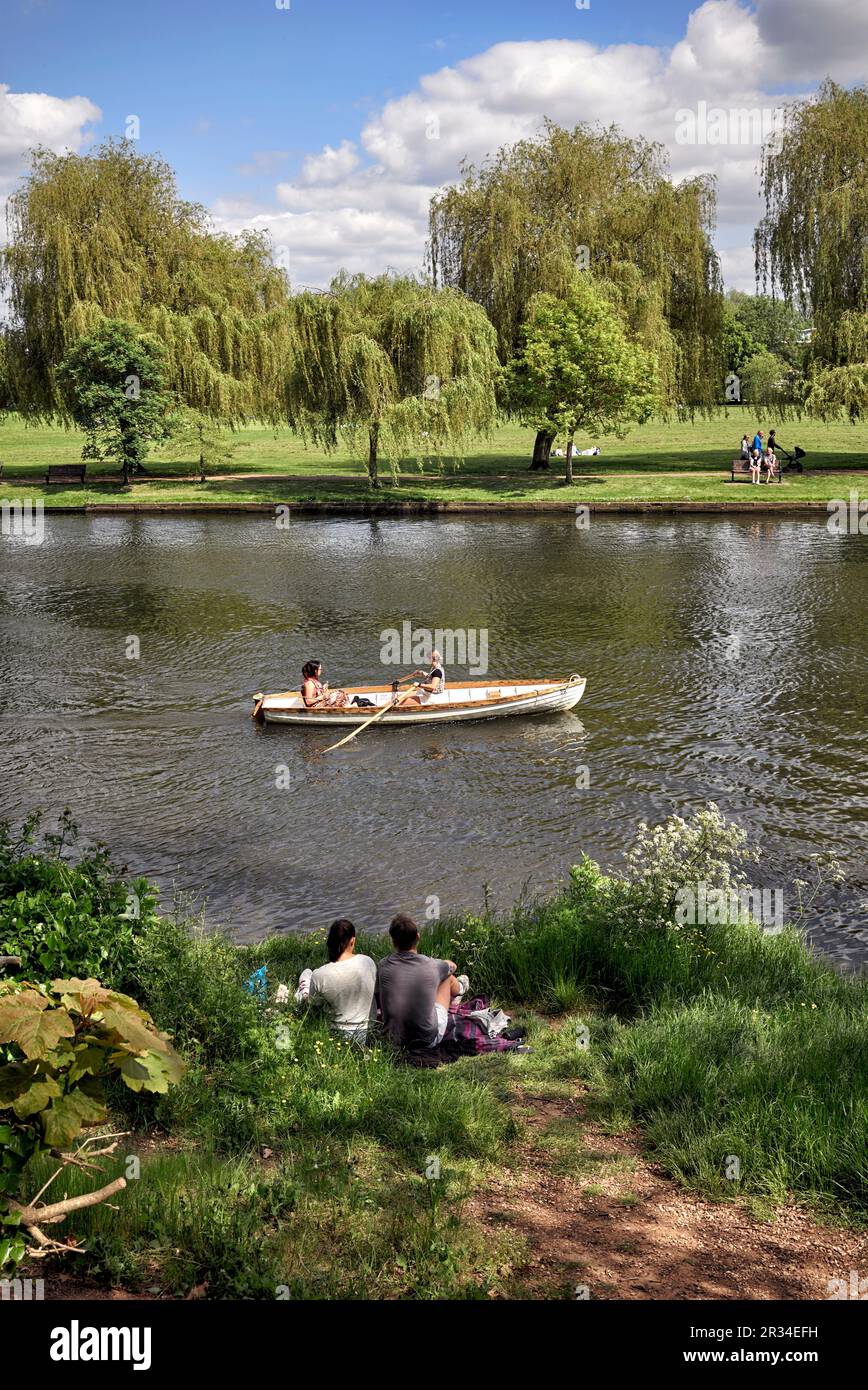 People sat relaxing at the riverside and others rowing boats on the ...