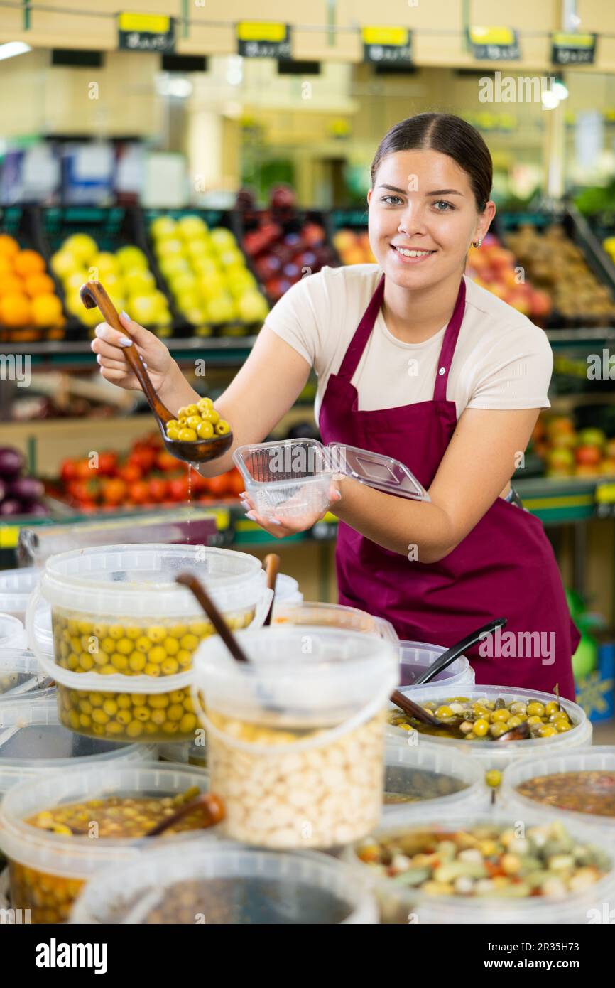 Positive female supermarket grocery clerk laying pickled olives in ...