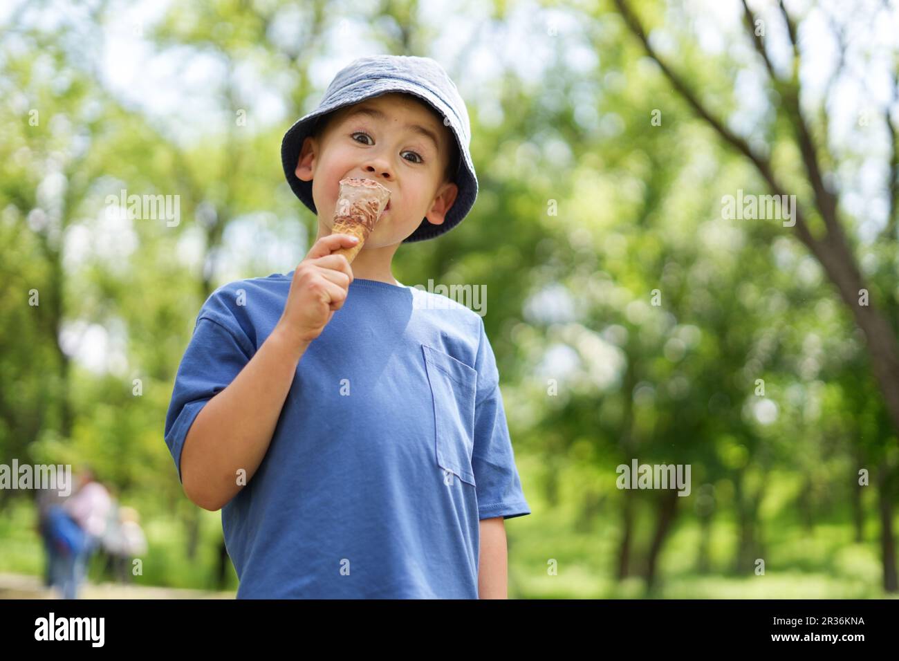 Cute little kid boy in a hat eating ice cream cone standing in a park ...