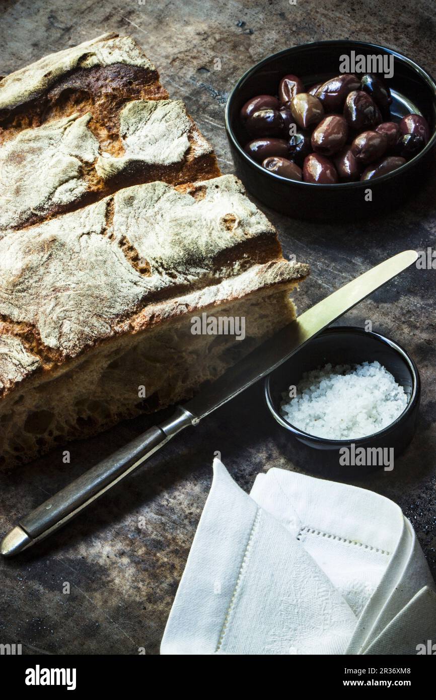 An arrangement of crusty bread, olives, sea salt, a knife and a fabric ...