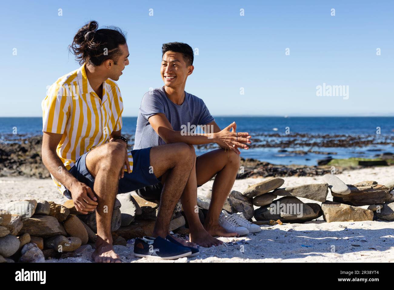 Happy biracial gay male couple sitting on rocks and talking on sunny ...