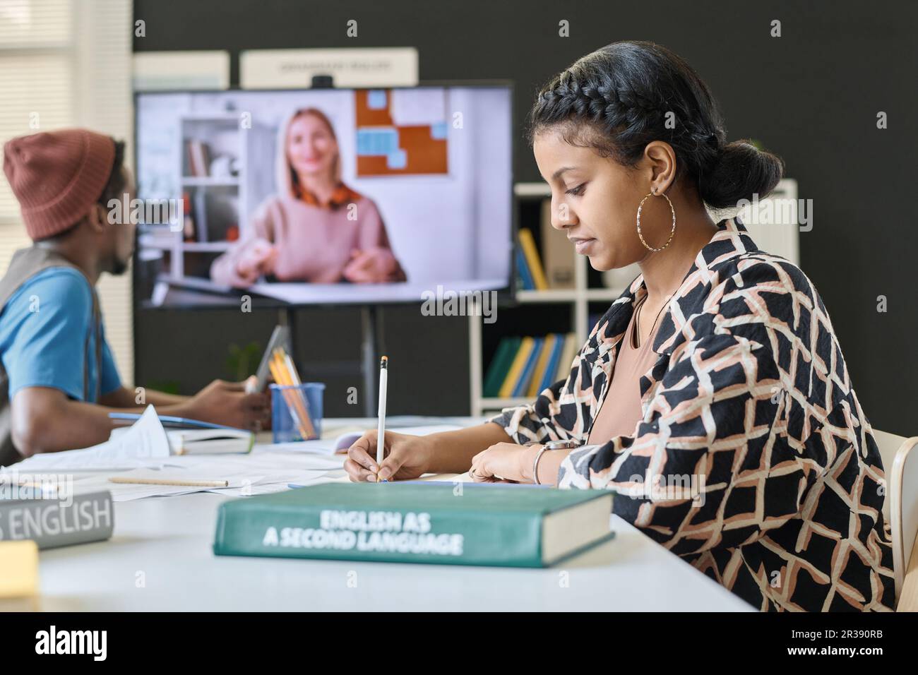 Student making notes at table while studying at lesson with teacher on ...