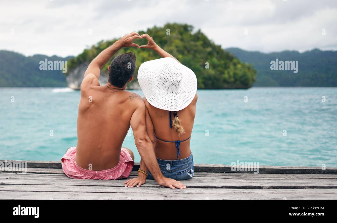 Heart hands, back and couple by a ocean while on summer, paradise and tropical vacation. Romance ...