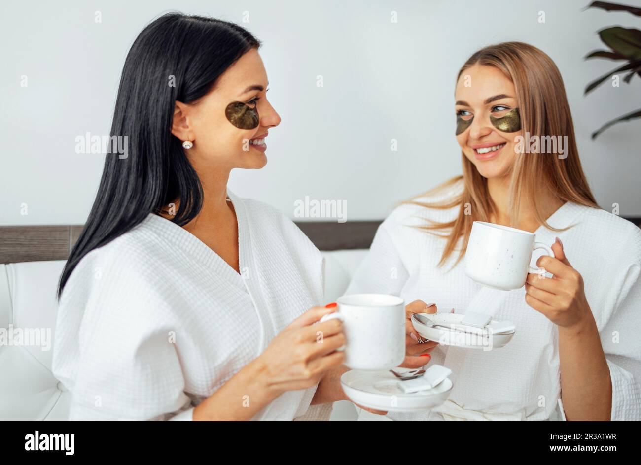 Two young female clients of beauty salon in white coats drink tea and ...
