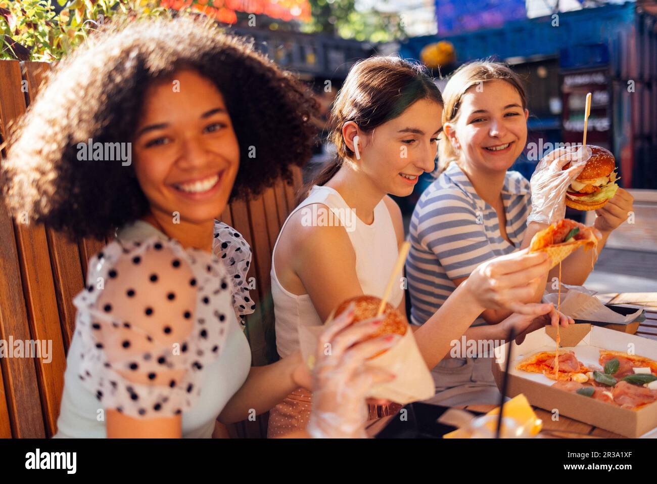 Cute smiling teenage girls are sitting in open air cafe and eating fast ...