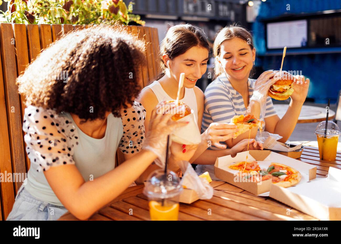 Cute smiling teenage girls are sitting in open air cafe and eating fast ...