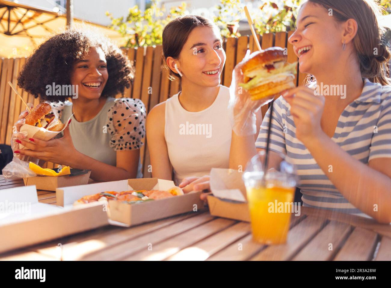 Cute smiling teenage girls are sitting in open air cafe and eating fast ...
