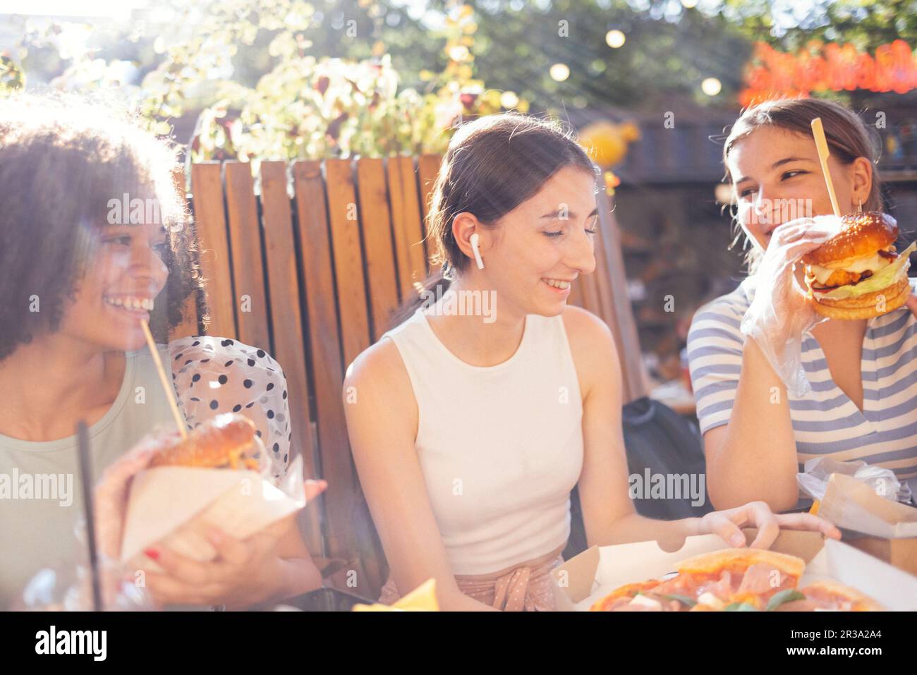 Cute smiling teenage girls are sitting in open air cafe and eating fast ...