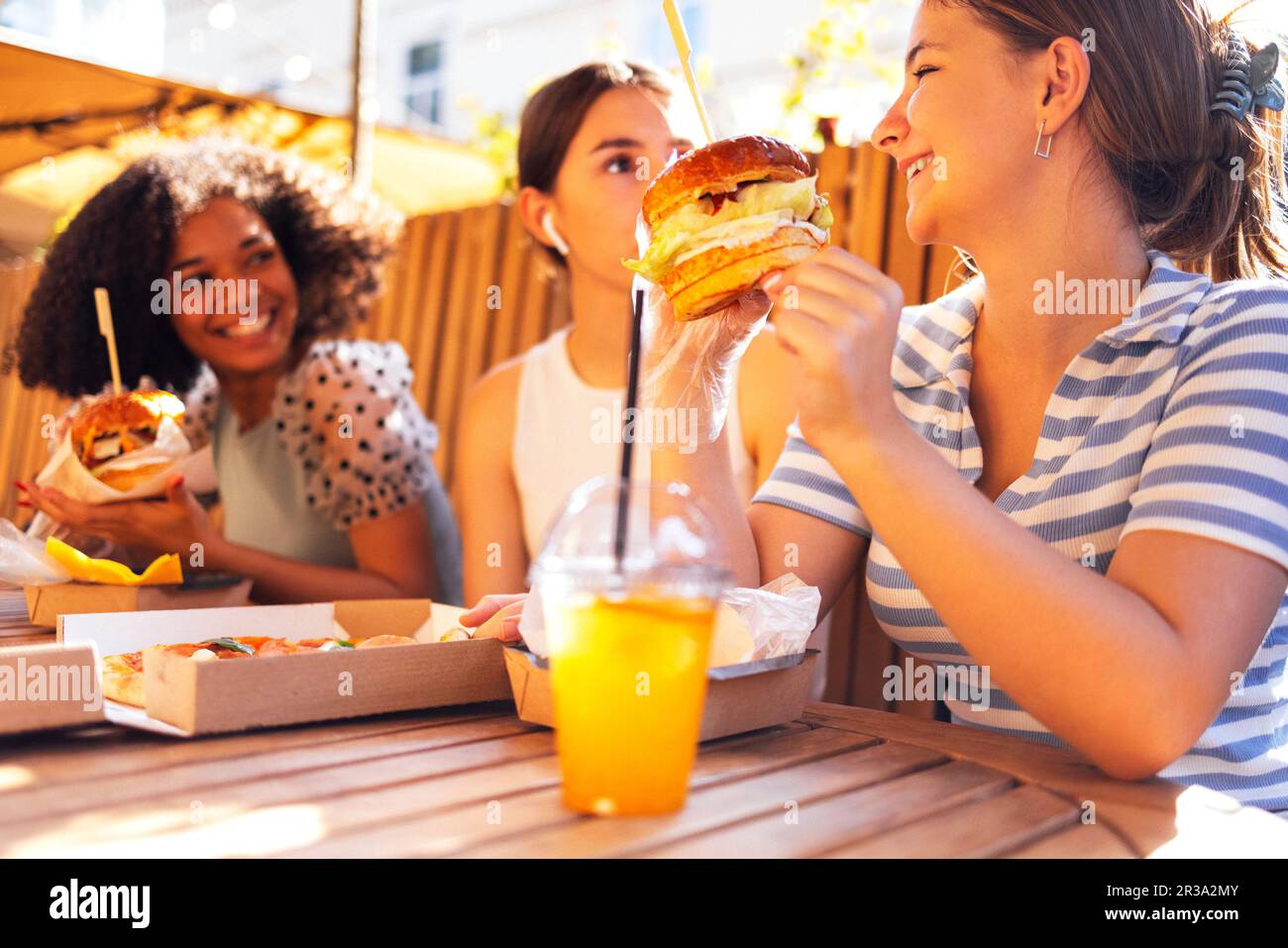 Cute smiling teenage girls are sitting in open air cafe and eating fast ...