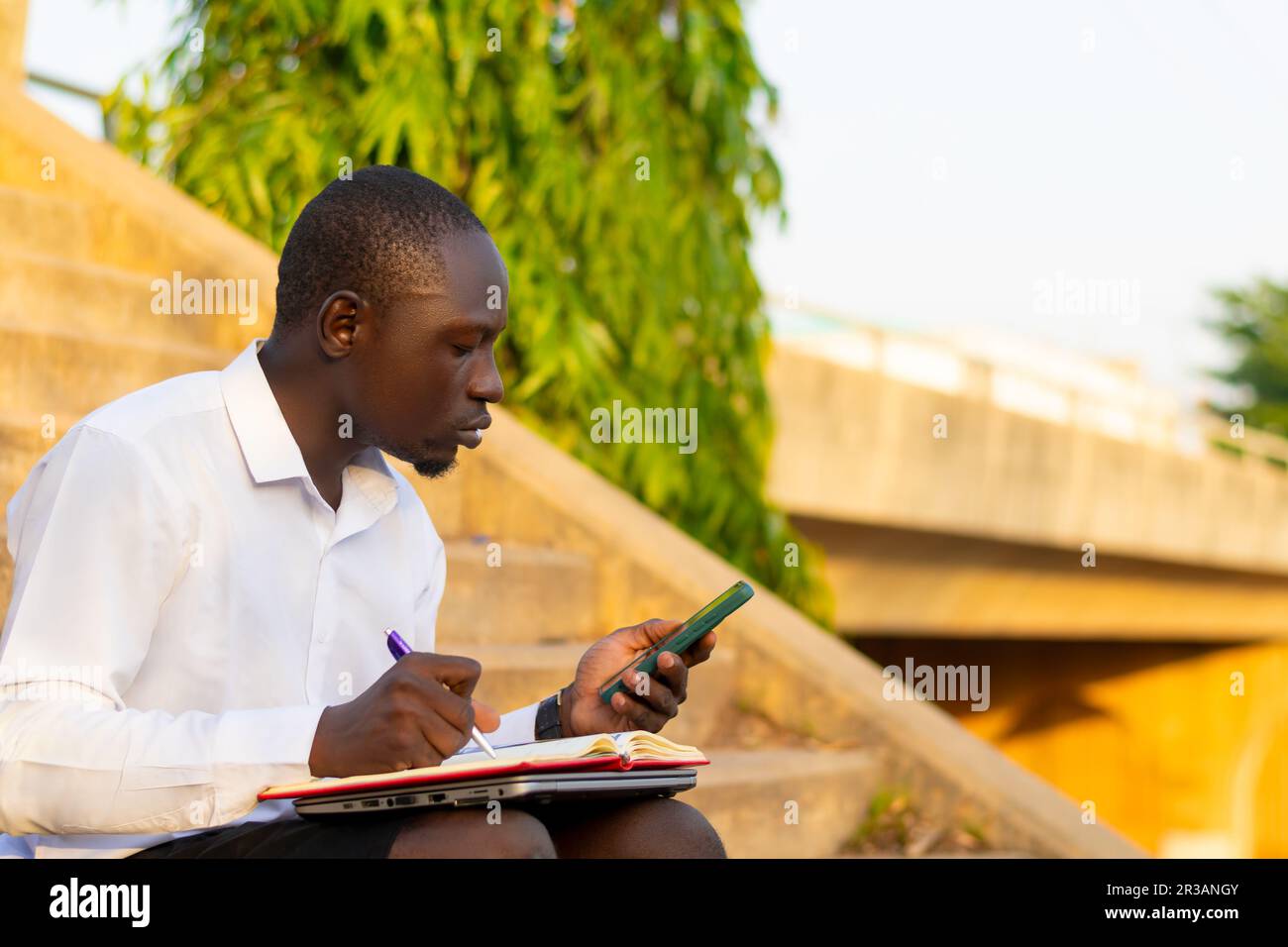 Business man seated writing on a notepad and using his mobile phone ...