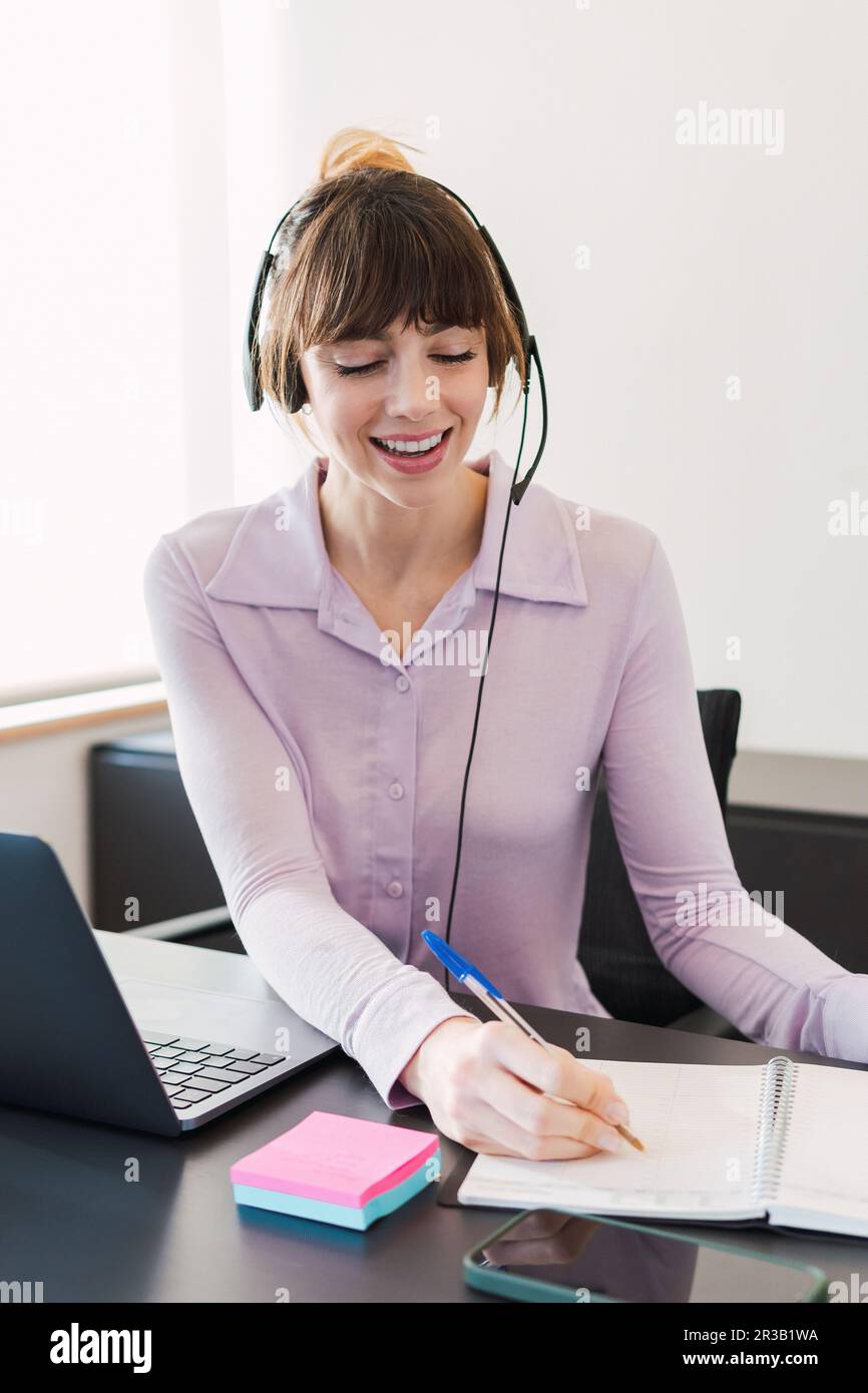 Happy businesswoman wearing headset writing on note pad at desk Stock ...