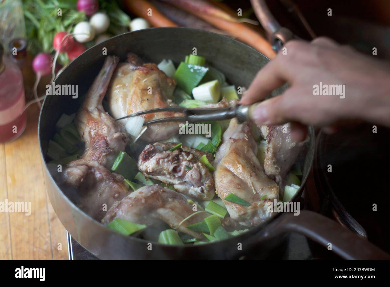 Hand holding tined fork stirs a copper pan of seared meat with leeks ...