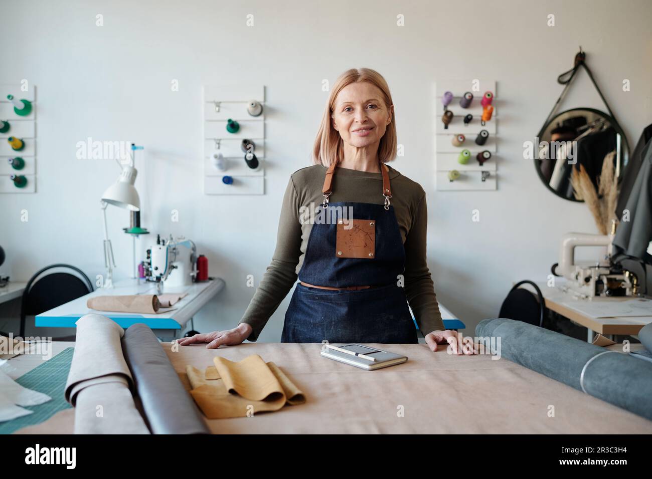 Smiling mature craftswoman in apron looking at camera in workshop or ...