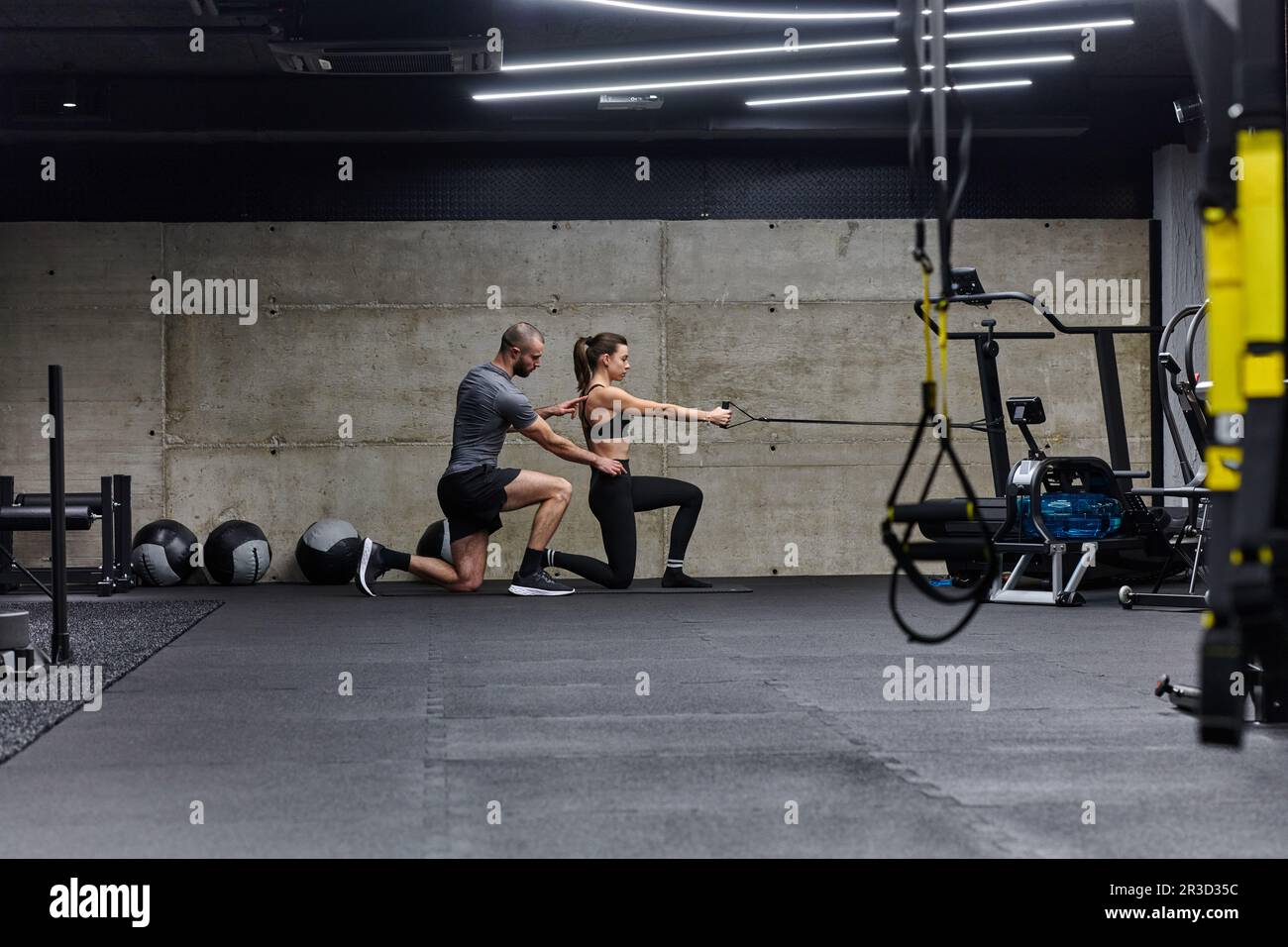 A muscular man assisting a fit woman in a modern gym as they engage in ...