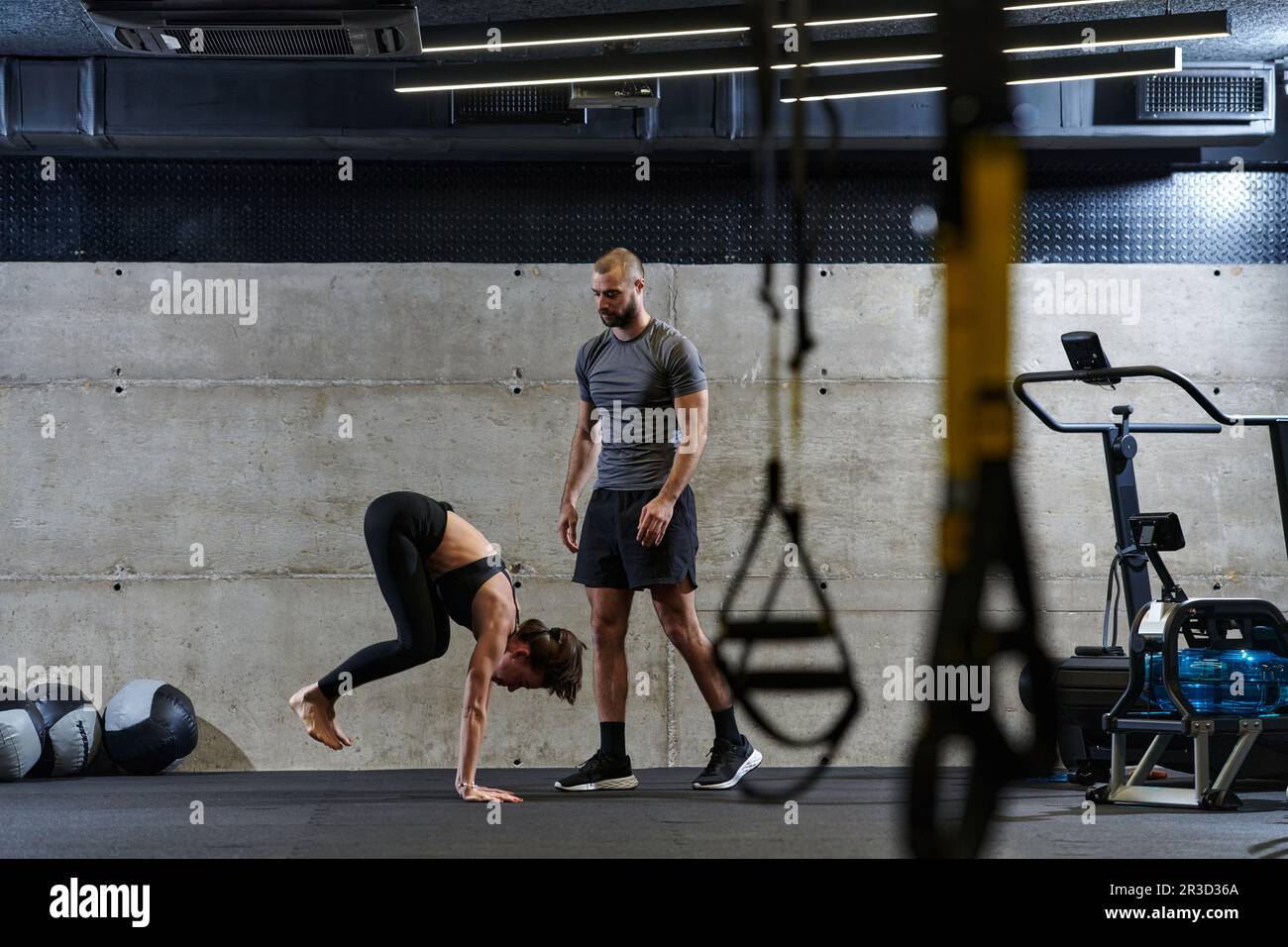 A muscular man assisting a fit woman in a modern gym as they engage in ...