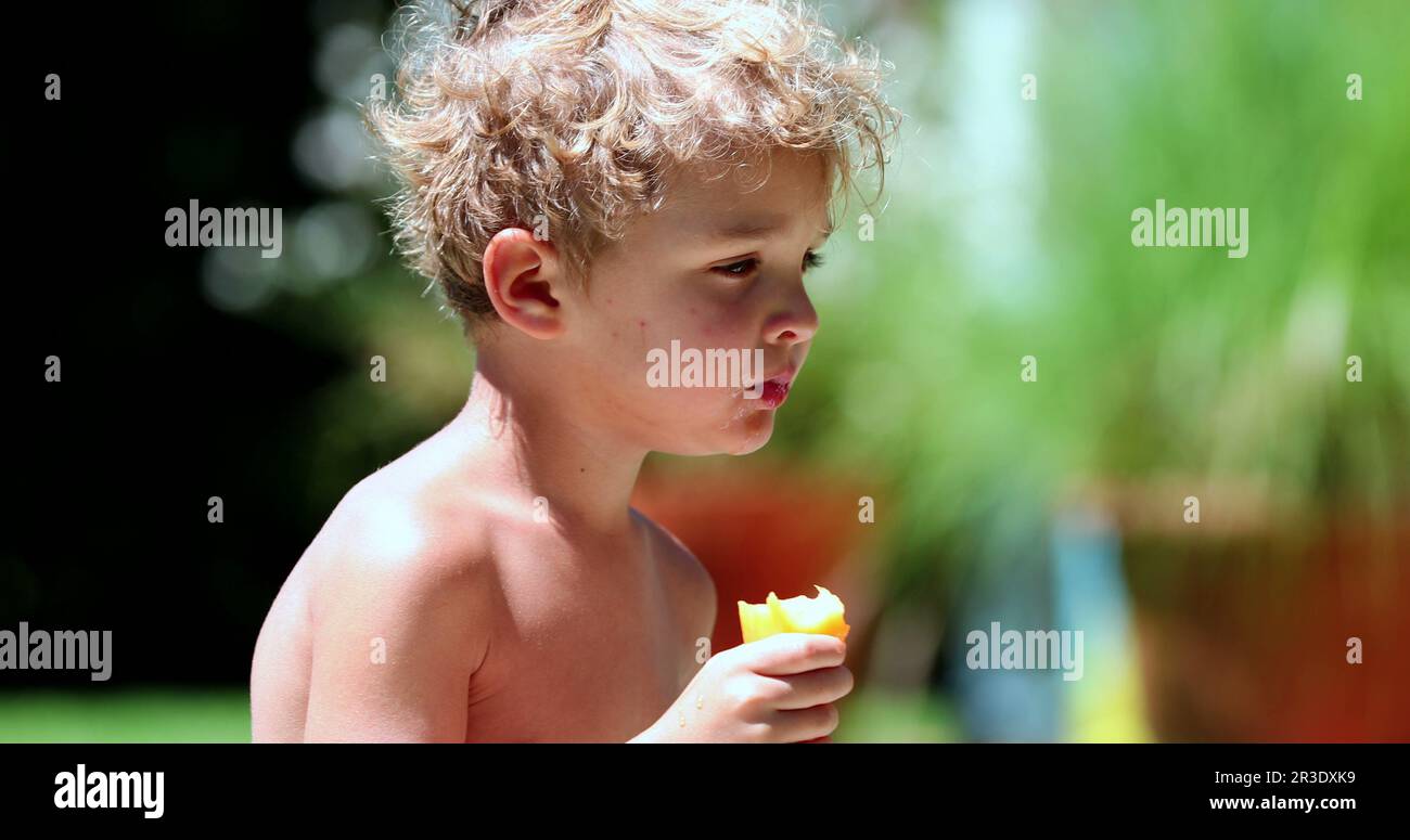 Cute toddler boy eating fruit snack outdoors in sunlight. Child chewing ...