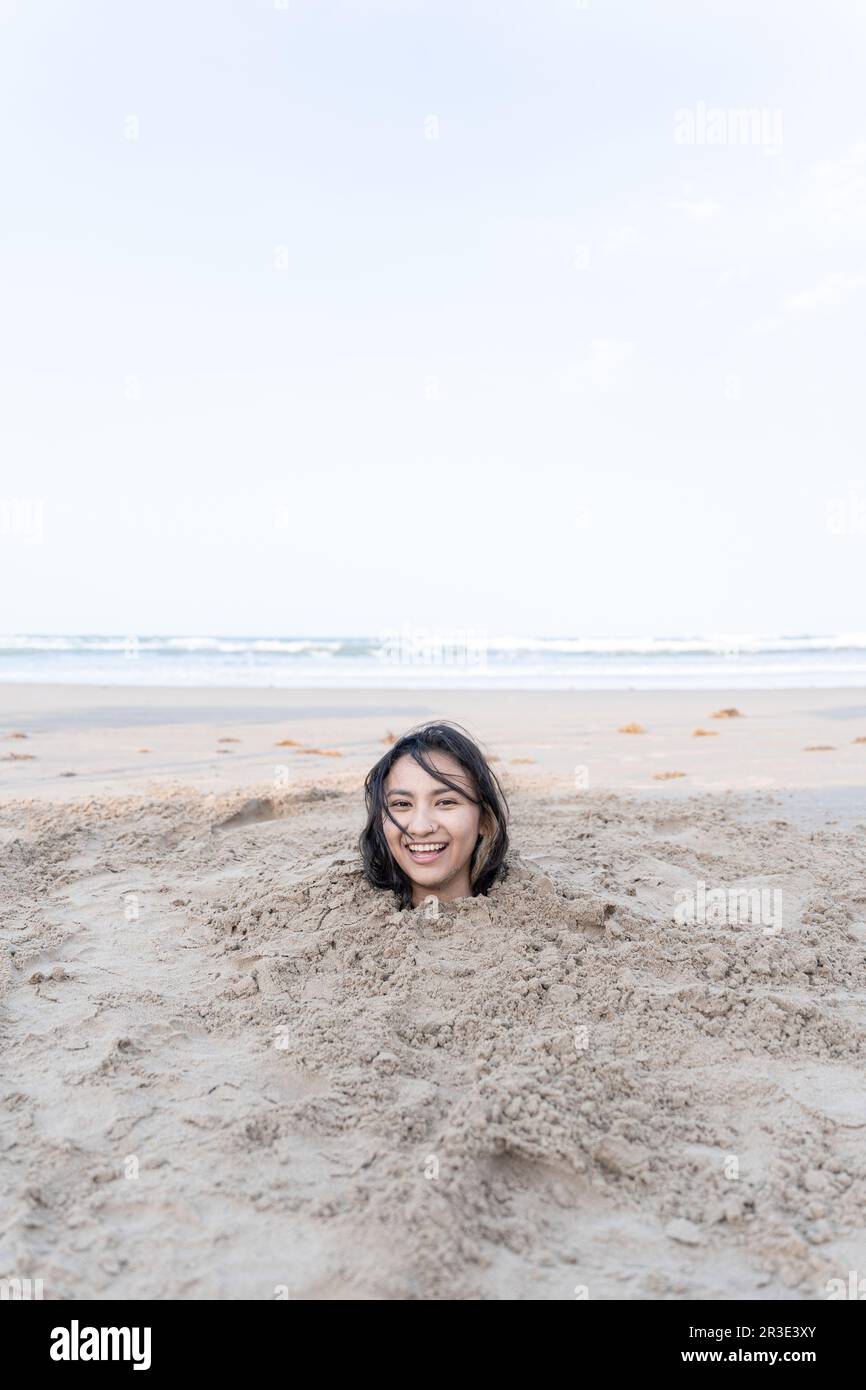A young Hispanic woman is having fun while buried in the sand of the ...
