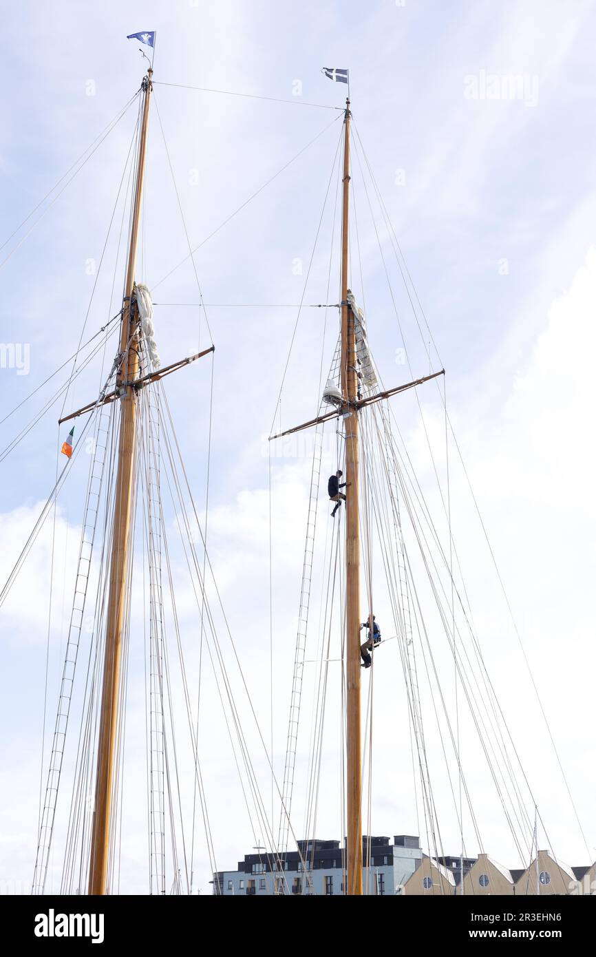 Two men dangling very high as they work on a tall mast of a boat in ...
