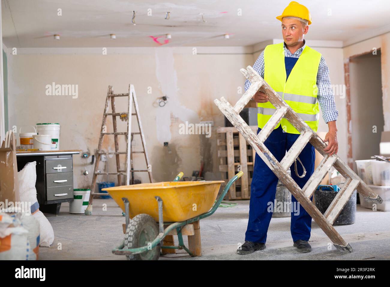 Worker in yellow vest carries a ladder on his shoulder in room being ...