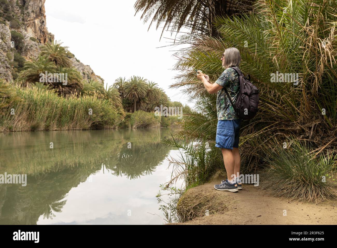 A gray-haired man with long hair stands on the bank of the river and ...