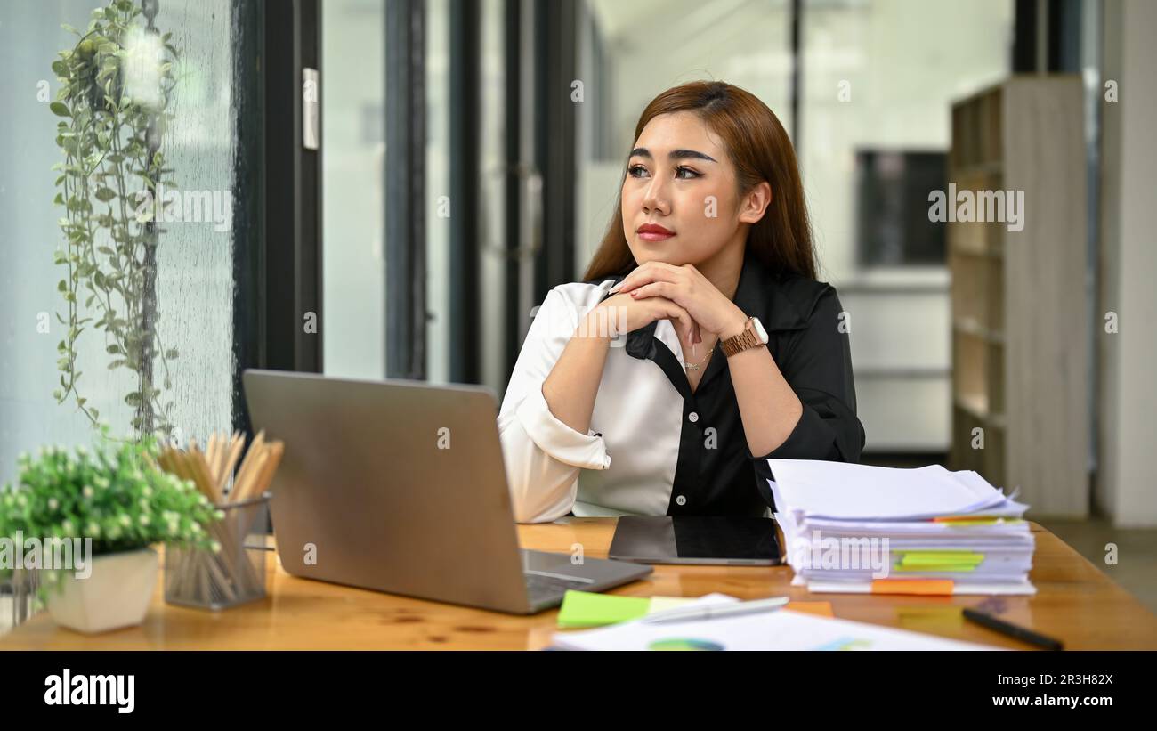 Thoughtful millennial Asian female accountant is looking out the window ...