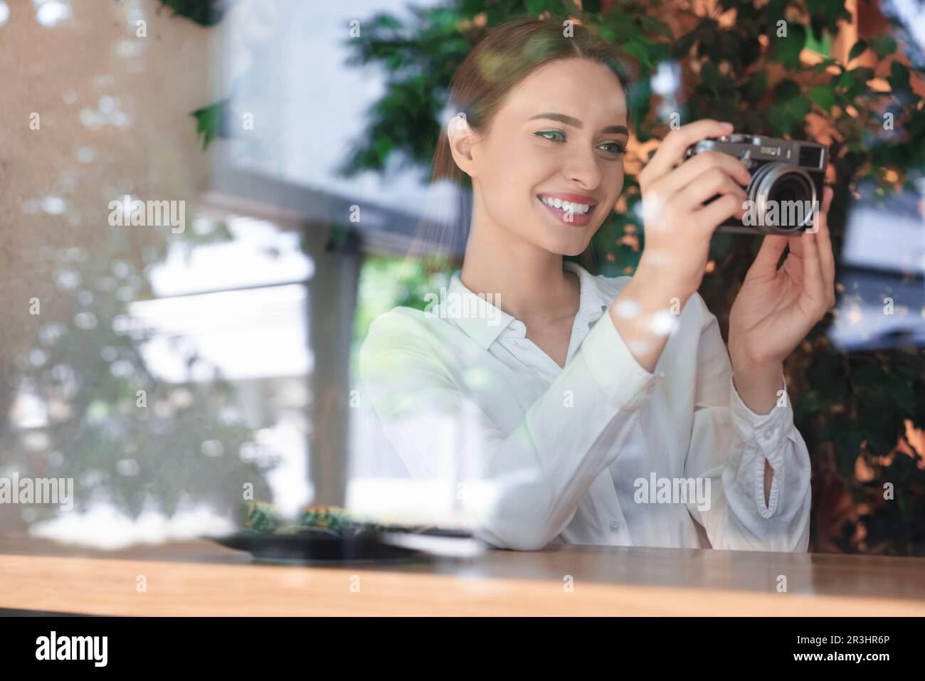 Young woman with camera taking photo at cafe, view through window ...