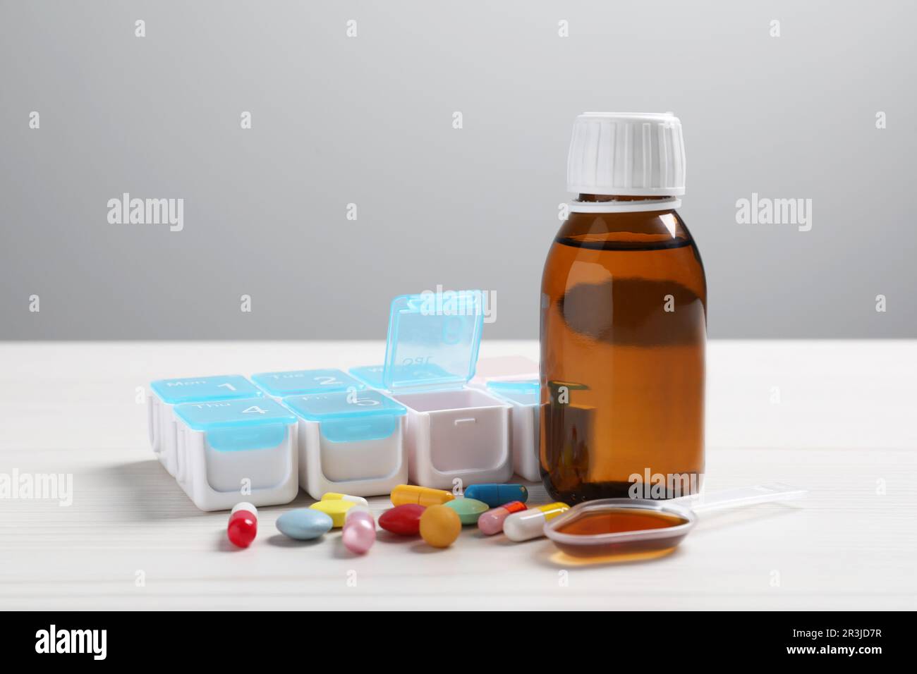 Bottle of syrup, dosing spoon and pills on white table against light ...