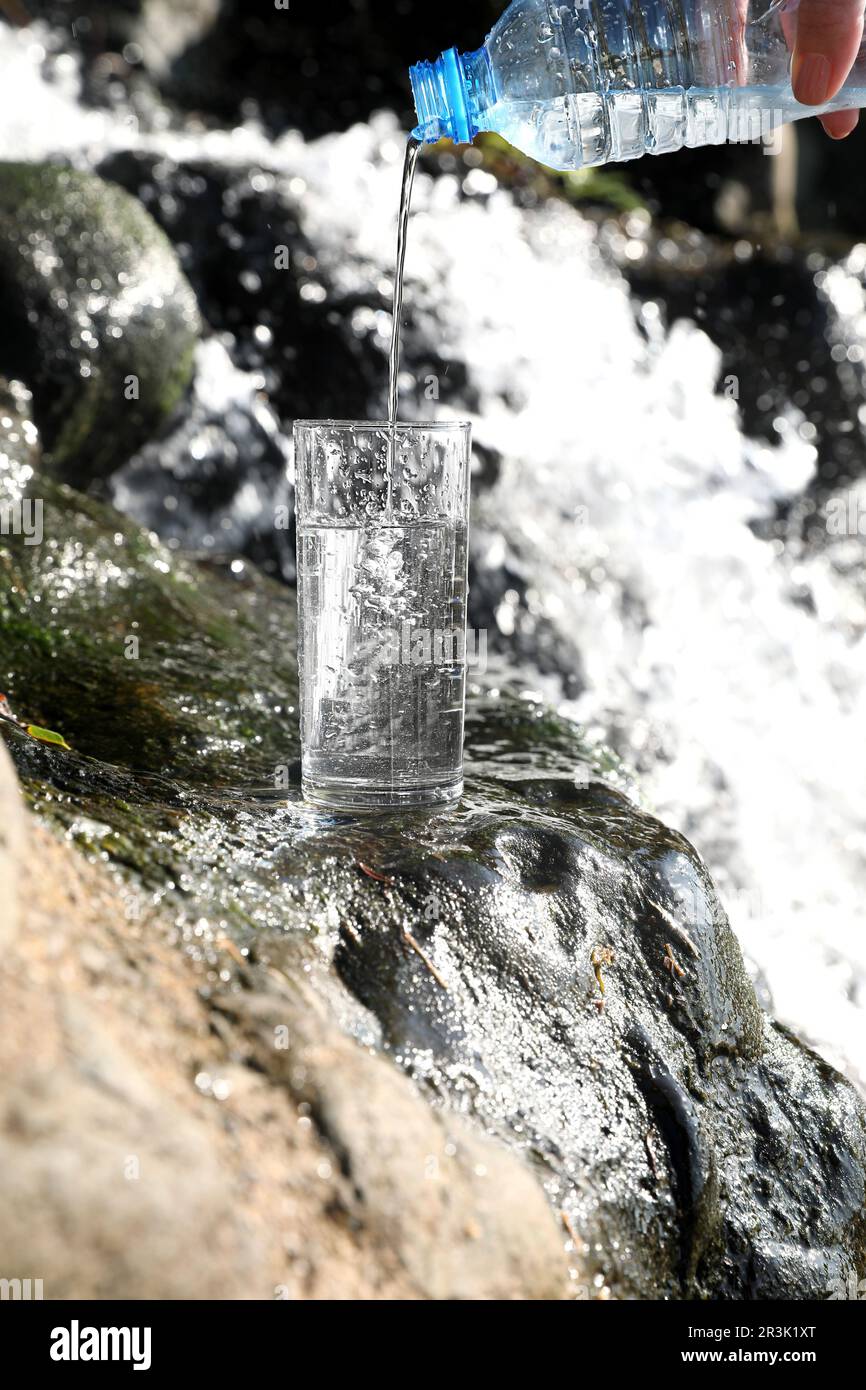 Woman pouring water from plastic bottle into glass near stream outdoors ...