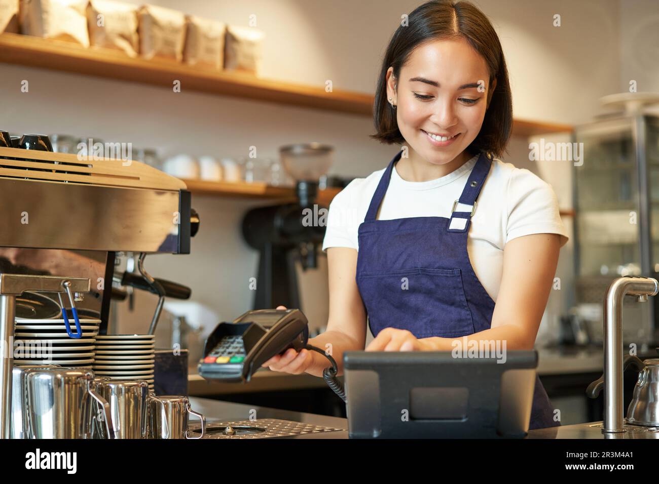 Smiling asian barista girl processing order, enter order in POS terminal, working at counter in ...