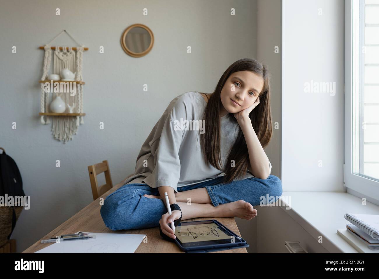 Positive serene teen girl sitting with legs crossed on table and ...