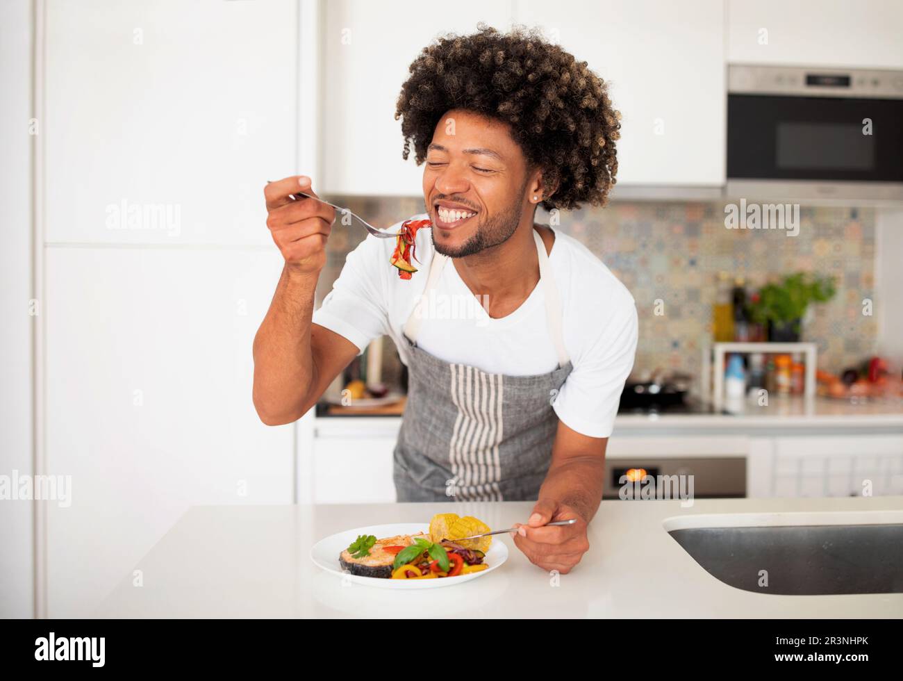 Cheerful black guy eating his homemade dinner at modern kitchen Stock ...