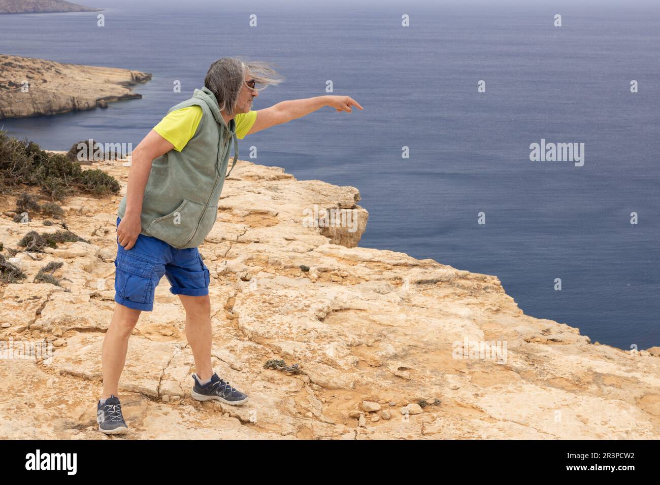 Gray-haired athletic man stands on a high cliff and points to the sea ...