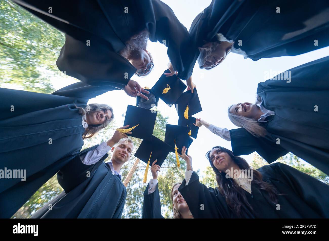 Classmates in graduation gowns toss their hats outdoors. Bottom view ...
