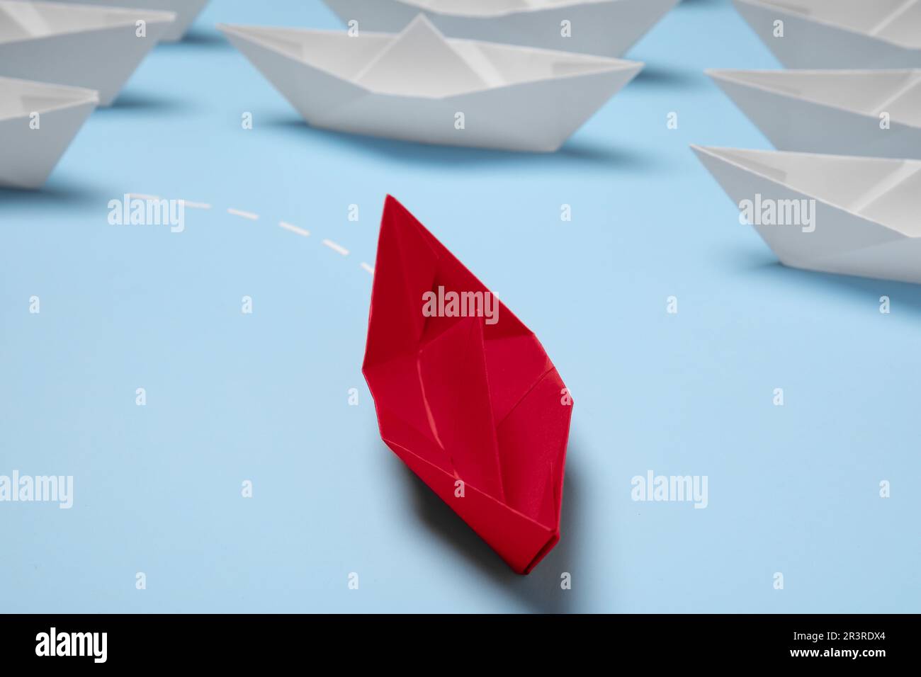 Red paper boat floating away from others on light blue background ...