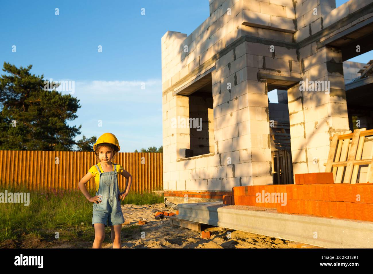 A little girl in a yellow hardhat is playing builder on the construction site of her future home ...