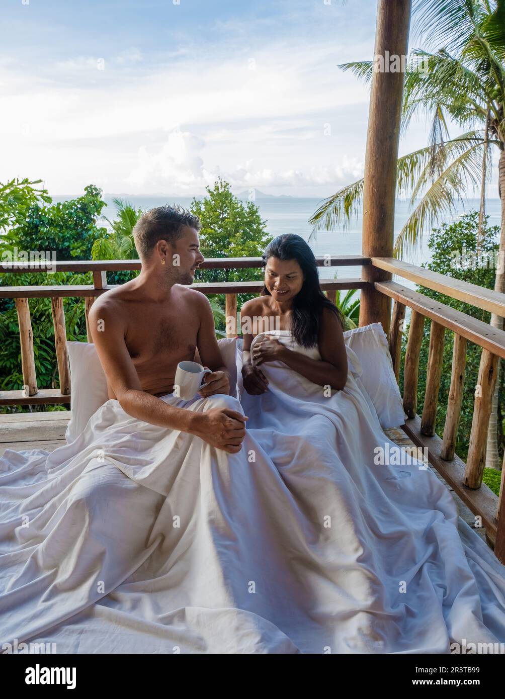 Couple waking up in bed looking out over ocean during sunrise at wooden ...