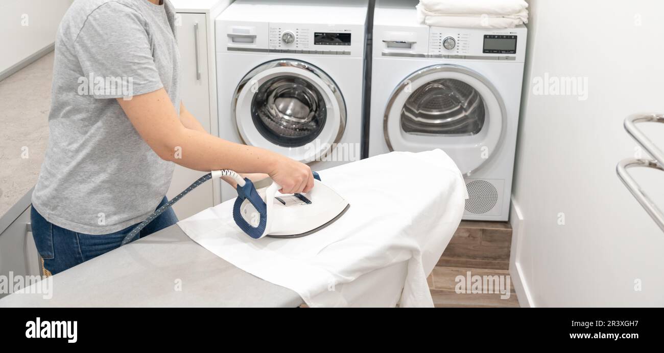 Woman ironing white shirt on board in laundry room with washing machine ...