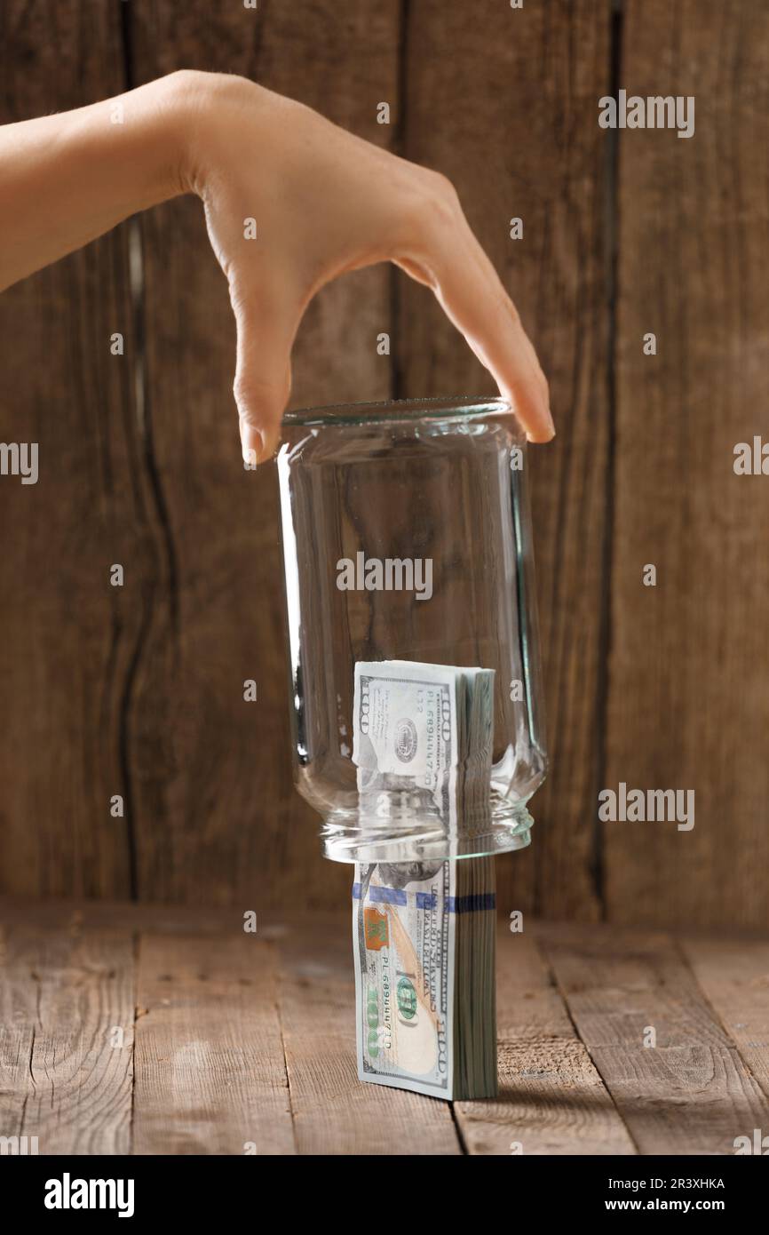 A hand raises a glass jar from which a stack of 100 dollar bills falls ...