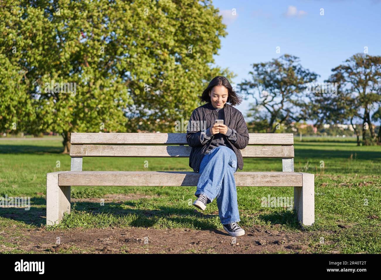 Young woman sitting alone on bench in park, using mobile phone, looking ...