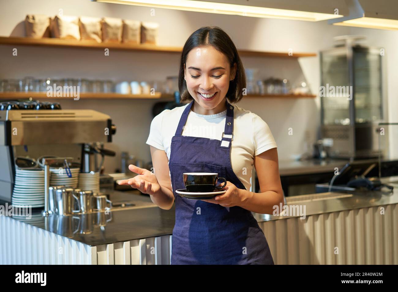 Portrait of smiling asian female barista, making coffee, holding cup of ...