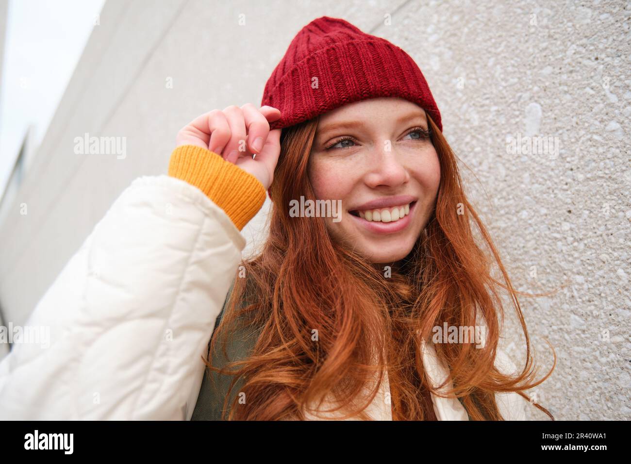 Stylish redhead girl in red hat, smiles and looks happy, poses outdoors ...